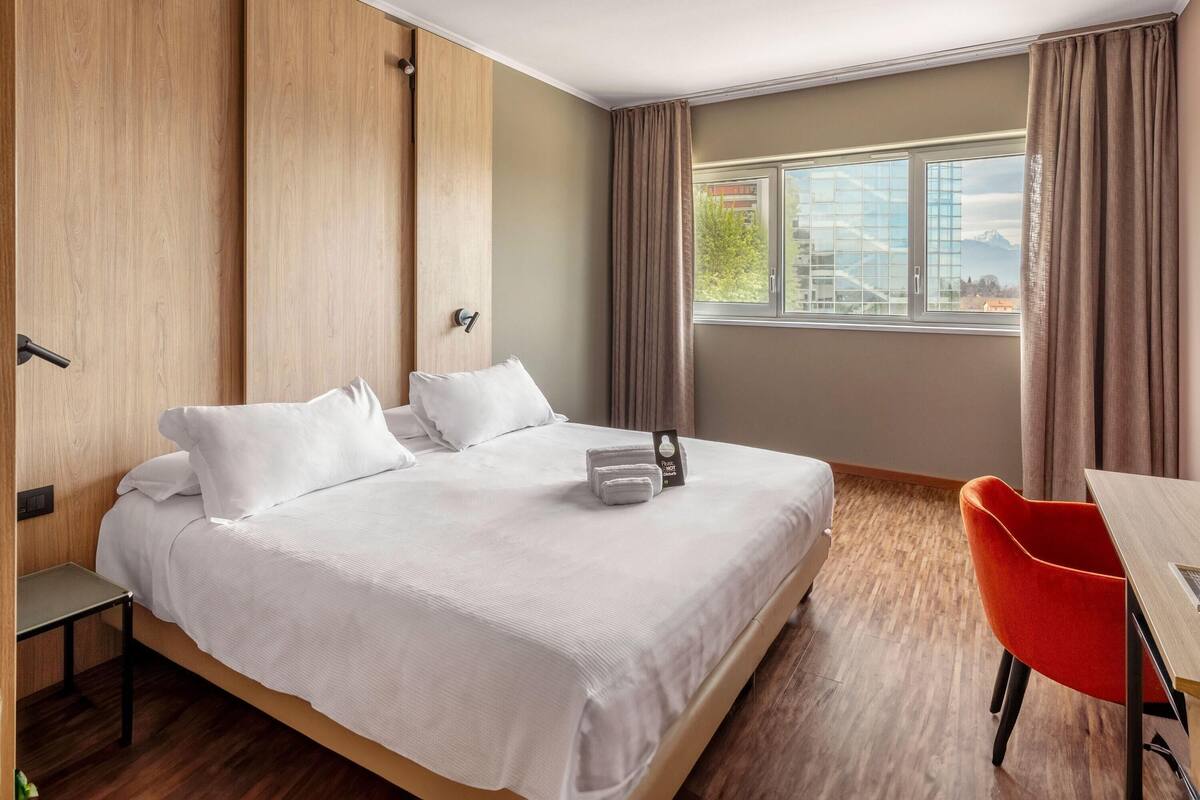 Quarto de hotel com uma cama grande, uma mesa e vista para a paisagem urbana pela janela.