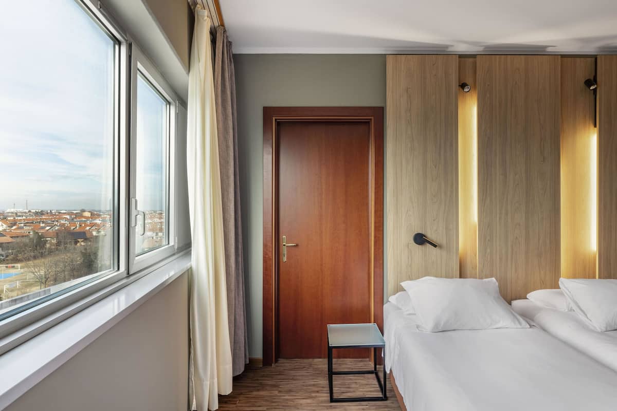 Quarto de hotel com uma janela grande, uma porta de madeira, uma cama com roupa de cama branca e uma pequena mesa.