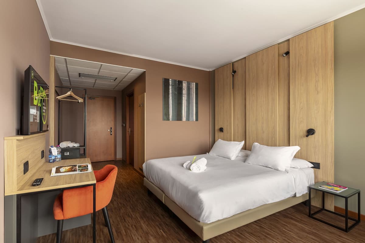 Quarto de hotel com uma cama, uma escrivaninha, uma cadeira laranja, uma mesinha de centro com uma revista e um guarda-roupa de madeira.
