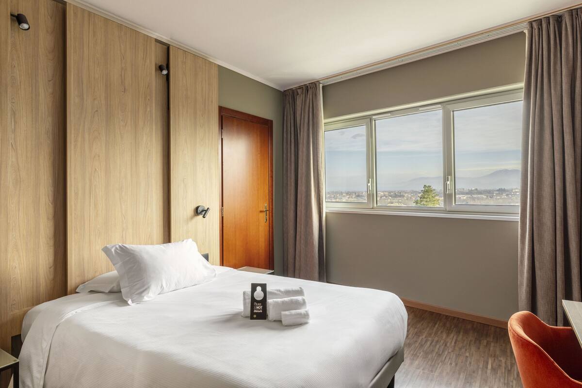 Quarto de hotel com uma cama, um guarda-roupa de madeira, uma janela com cortinas e vista para a paisagem urbana.