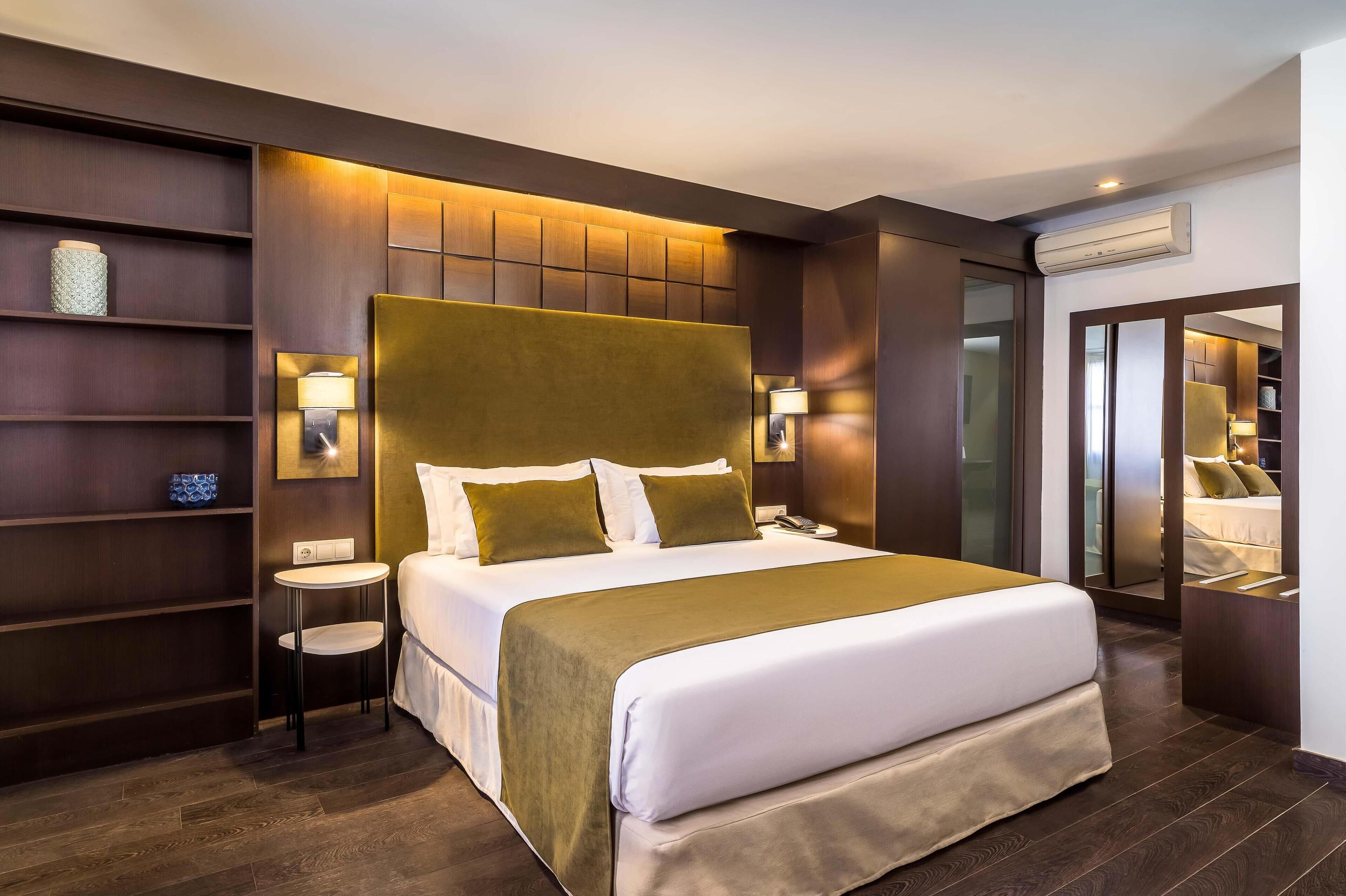 Quarto de hotel moderno com uma cama grande, estantes embutidas, abajures e um espelho.