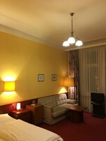 Quarto de hotel com cama, sofá, escrivaninha, televisão e lustre.