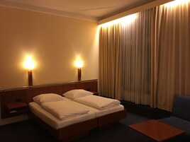 Quarto de hotel com cama de casal, duas luminárias de cabeceira, uma cadeira e uma janela com cortinas.
