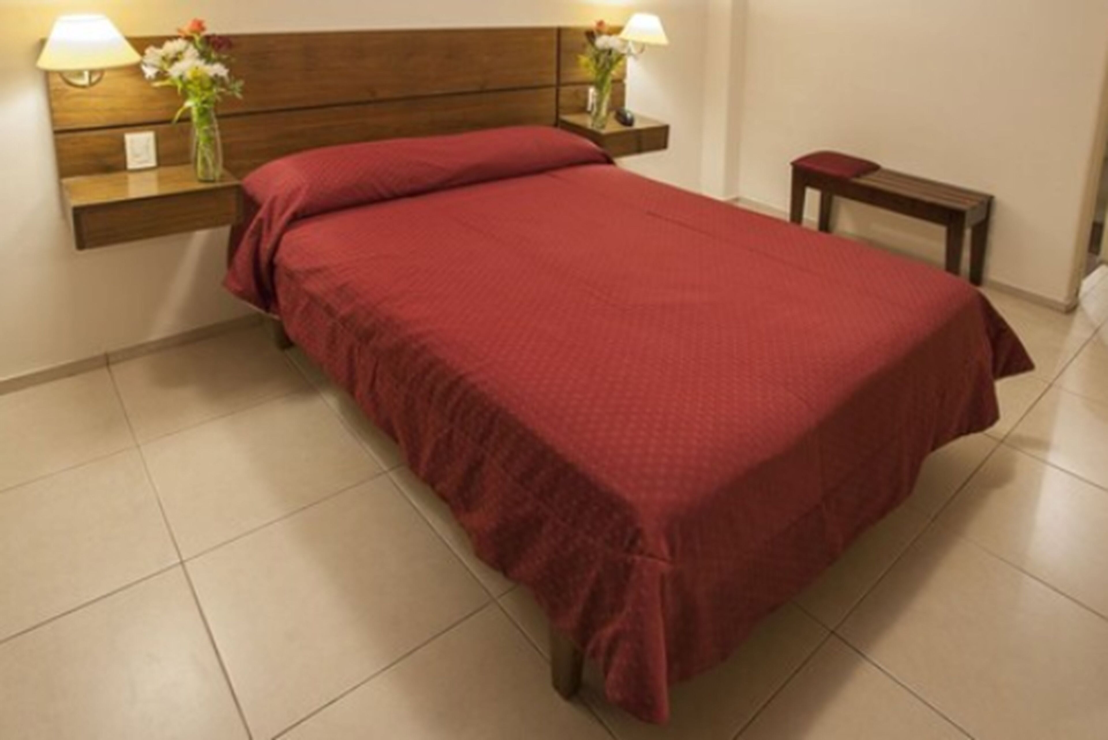Uma cama bem arrumada com um cobreleto vermelho, duas mesas de cabeceira com abajures e um pequeno banco.