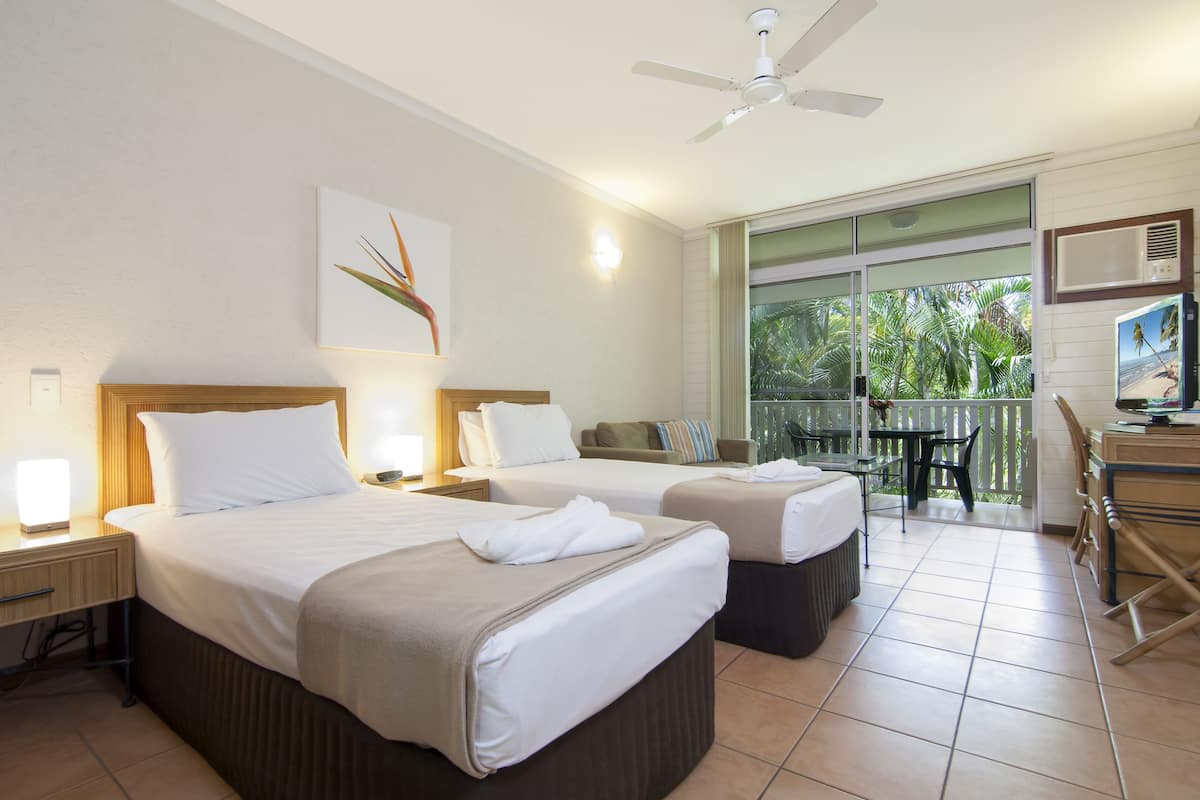 Quarto de hotel com duas camas, varanda com mesa e cadeiras, e um ventilador de teto.
