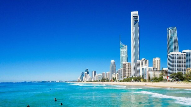Paisagem costeira com edifícios altos, águas azuis cristalinas e surfistas no oceano.