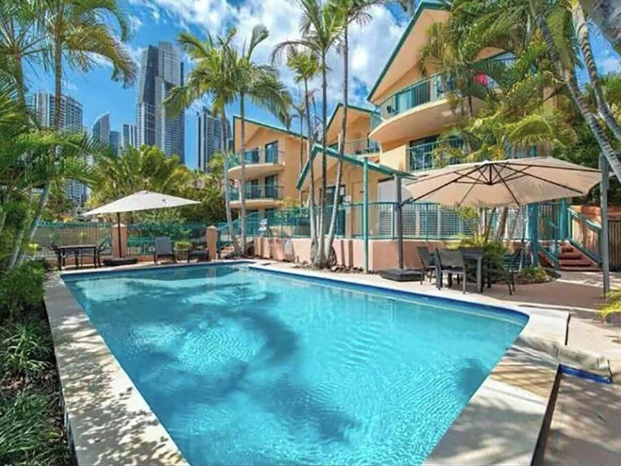 Piscina com superfície de água azul cristalina, rodeada por palmeiras e um edifício com varandas.