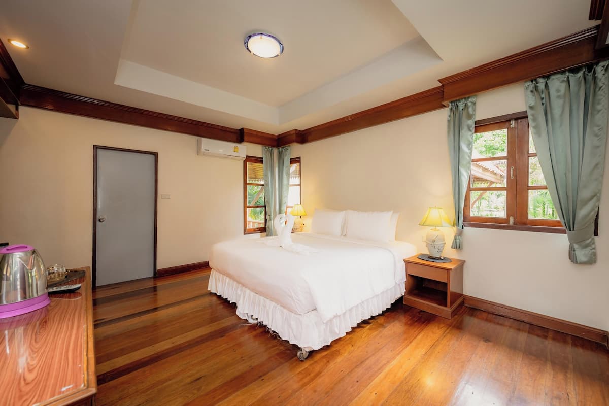 Quarto de hotel com chão de madeira, uma cama grande com roupa de cama branca, duas mesas de cabeceira com abajures, uma janela com cortinas e um aparelho de ar condicionado na parede.