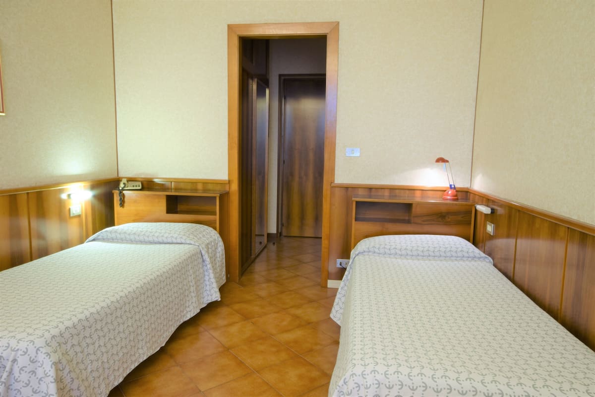 Quarto de hotel com duas camas de solteiro, cabeceiras de madeira, mesa de cabeceira com abajur e porta para outro quarto.