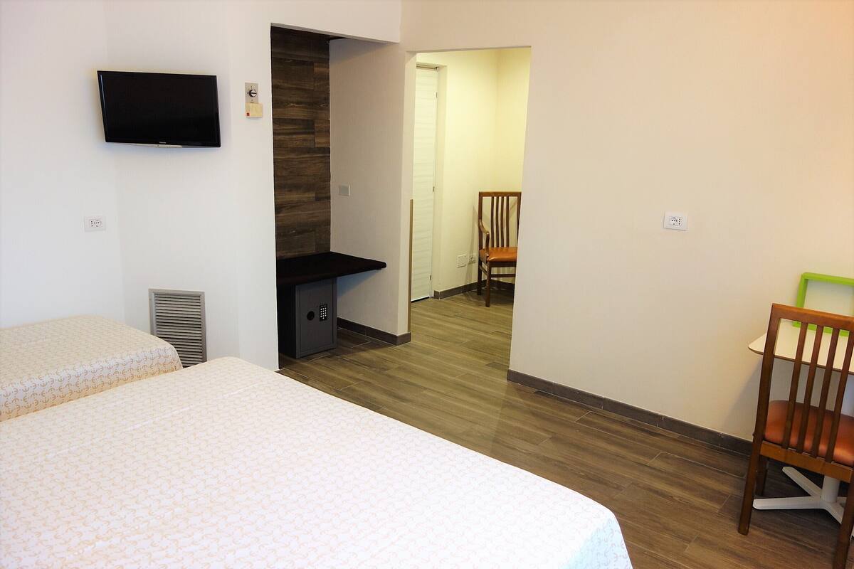 Quarto de hotel com duas camas, televisão na parede, piso de madeira e uma cadeira.