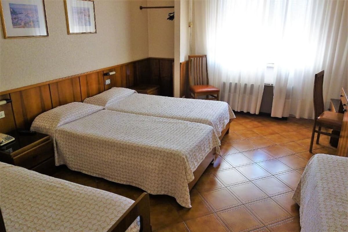 Quarto de hotel com duas camas, cabeceira de madeira, uma cadeira, uma mesinha e uma janela com cortinas.