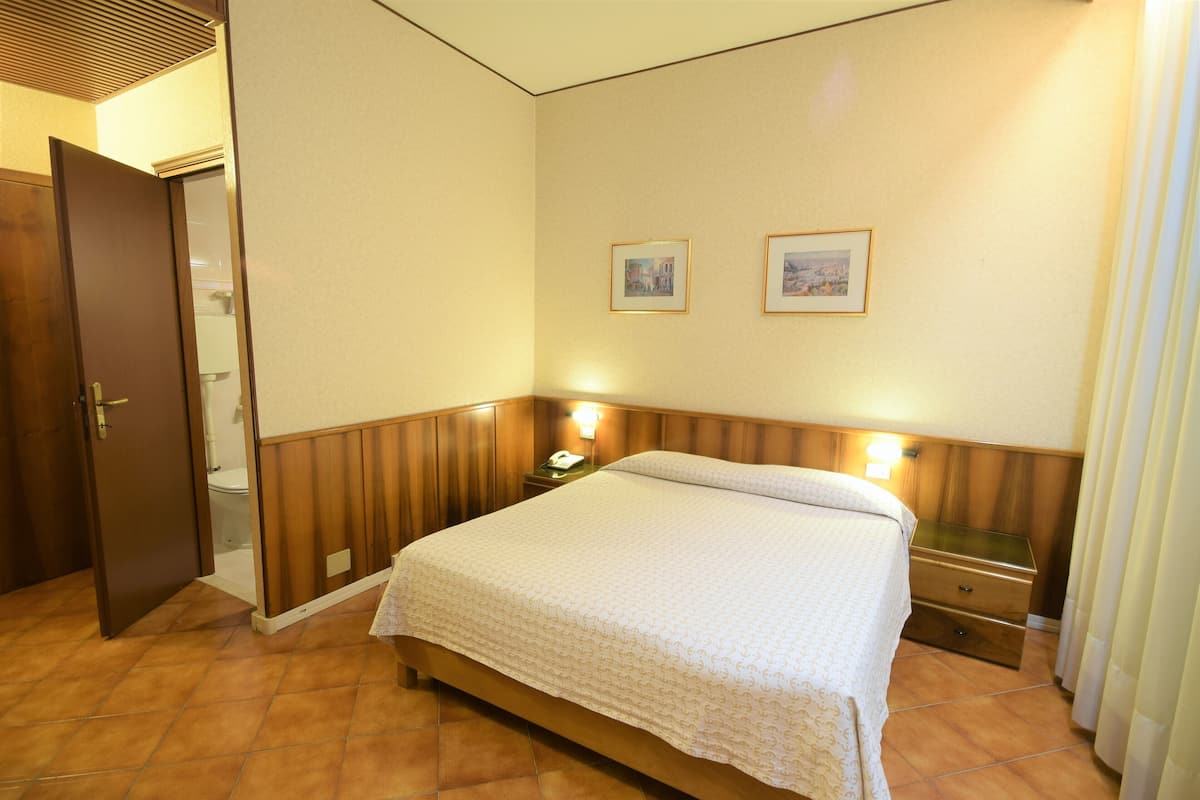 Quarto de hotel com uma cama, cabeceira de madeira, duas luminárias na parede, mesa de cabeceira com um telefone e uma porta para um banheiro.