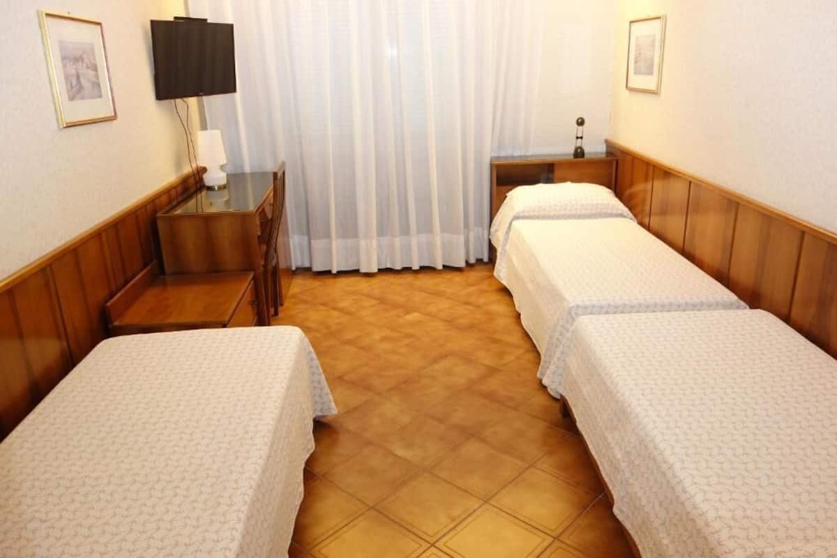 Quarto de hotel com duas camas de solteiro, uma cômoda de madeira, uma escrivaninha com abajur, televisão e uma janela com cortinas brancas.