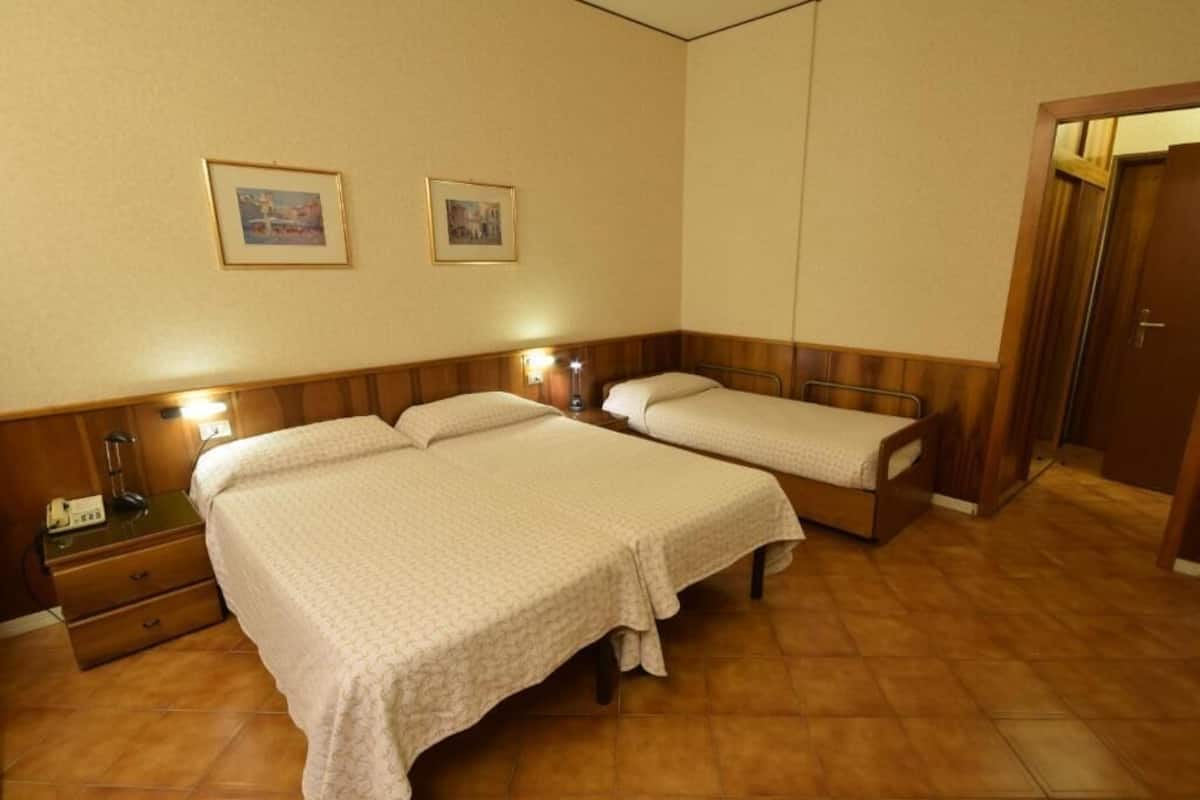 Quarto de hotel com duas camas, uma mesa de cabeceira com abajur e uma porta de madeira.