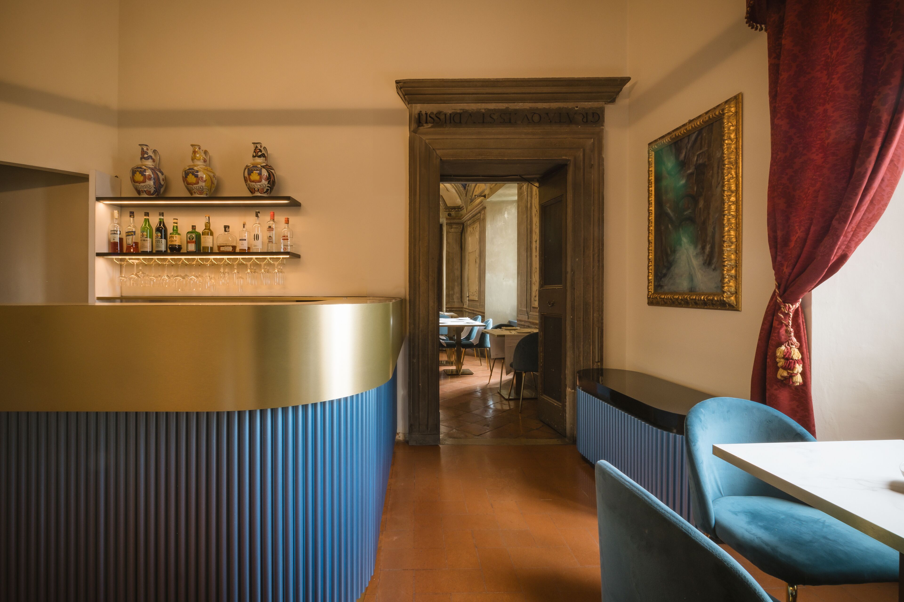 Uma área de bar com prateleiras de garrafas e copos, um balcão e um espaço de refeições com cadeiras azuis e uma mesa.