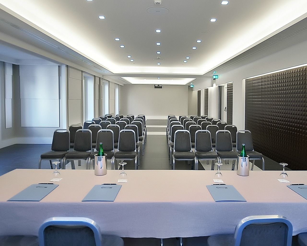 Uma sala de conferências com fileiras de cadeiras, uma mesa comprida e uma tela de projeção.
