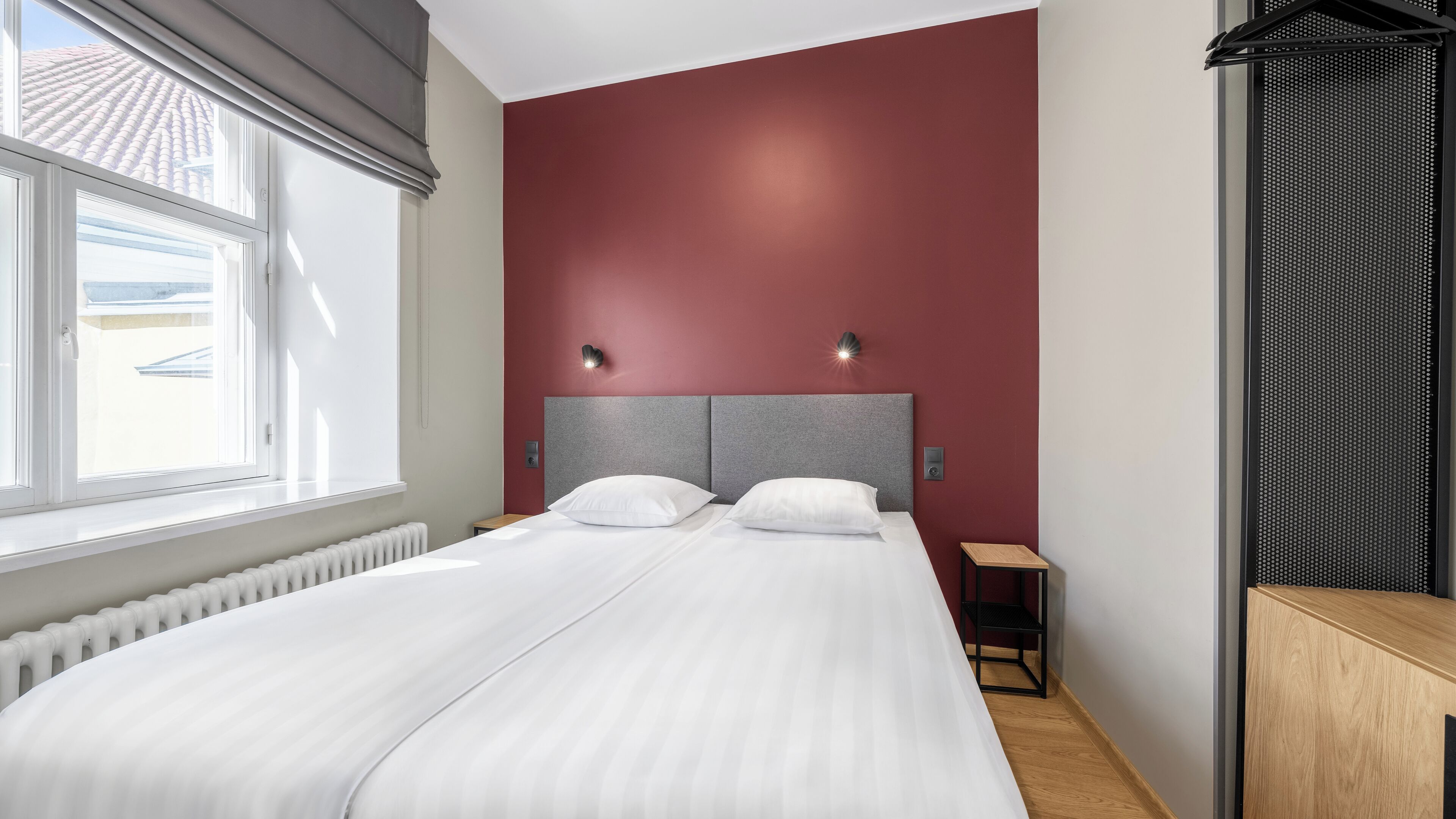 Quarto de hotel moderno com uma cama grande, um radiador, uma mesinha de madeira e uma prateleira fixada na parede.