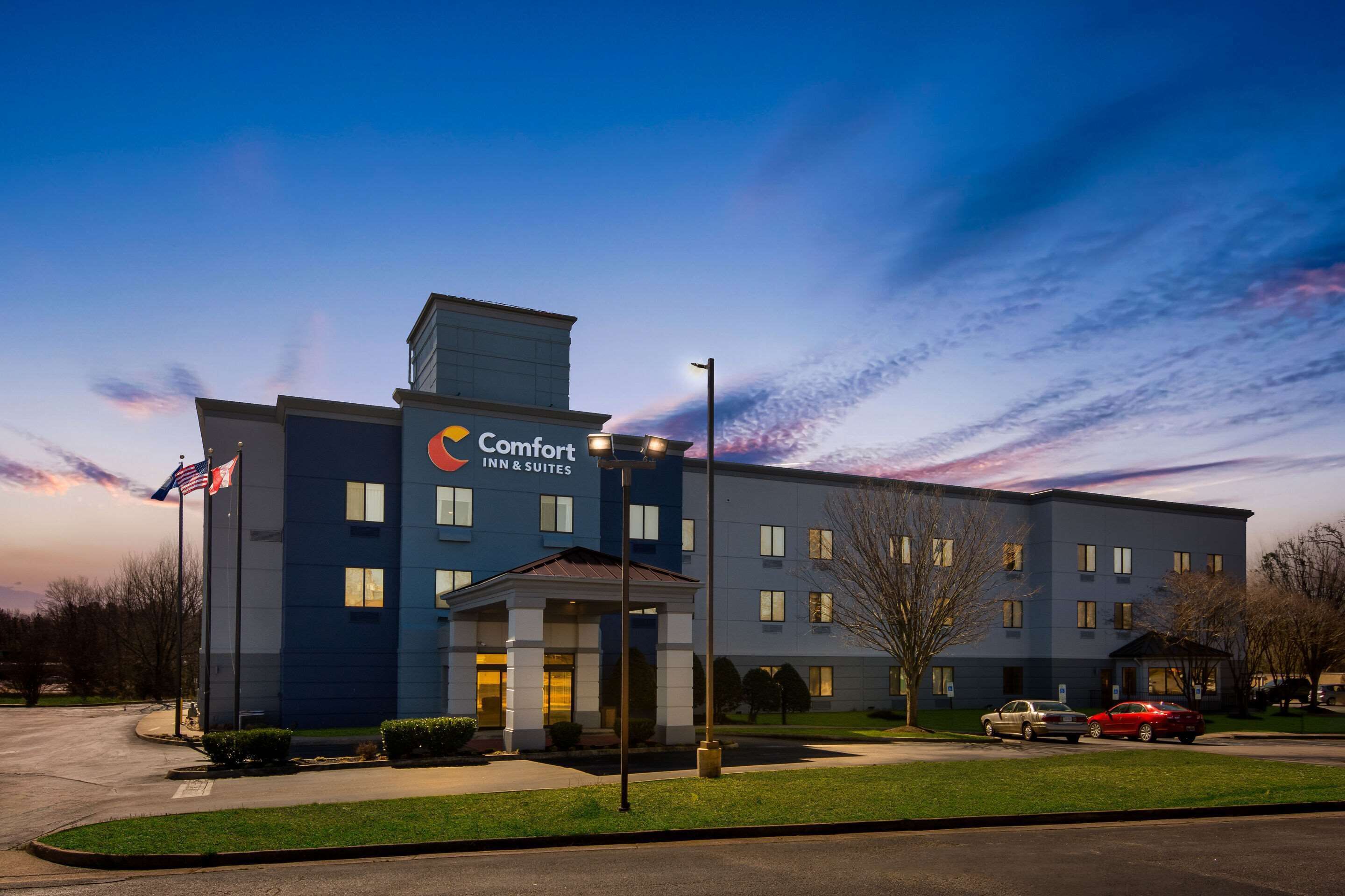 Um hotel Comfort Inn & Suites com design moderno, exibindo uma bandeira americana em destaque, uma entrada bem iluminada e um estacionamento com vários carros.