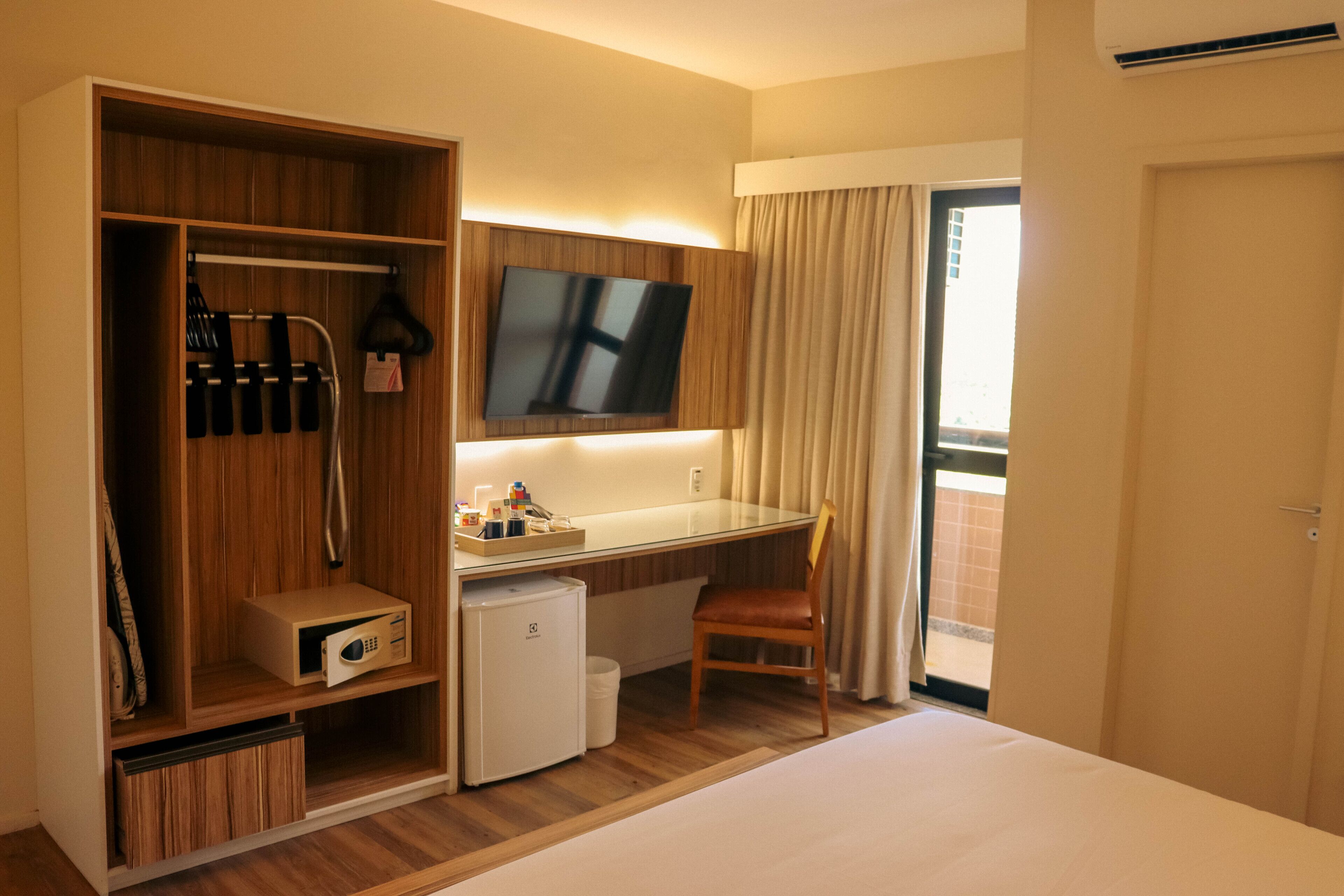 Quarto de hotel com uma cama, escrivaninha com televisão, guarda-roupa, pequeno frigorífico e uma cadeira.