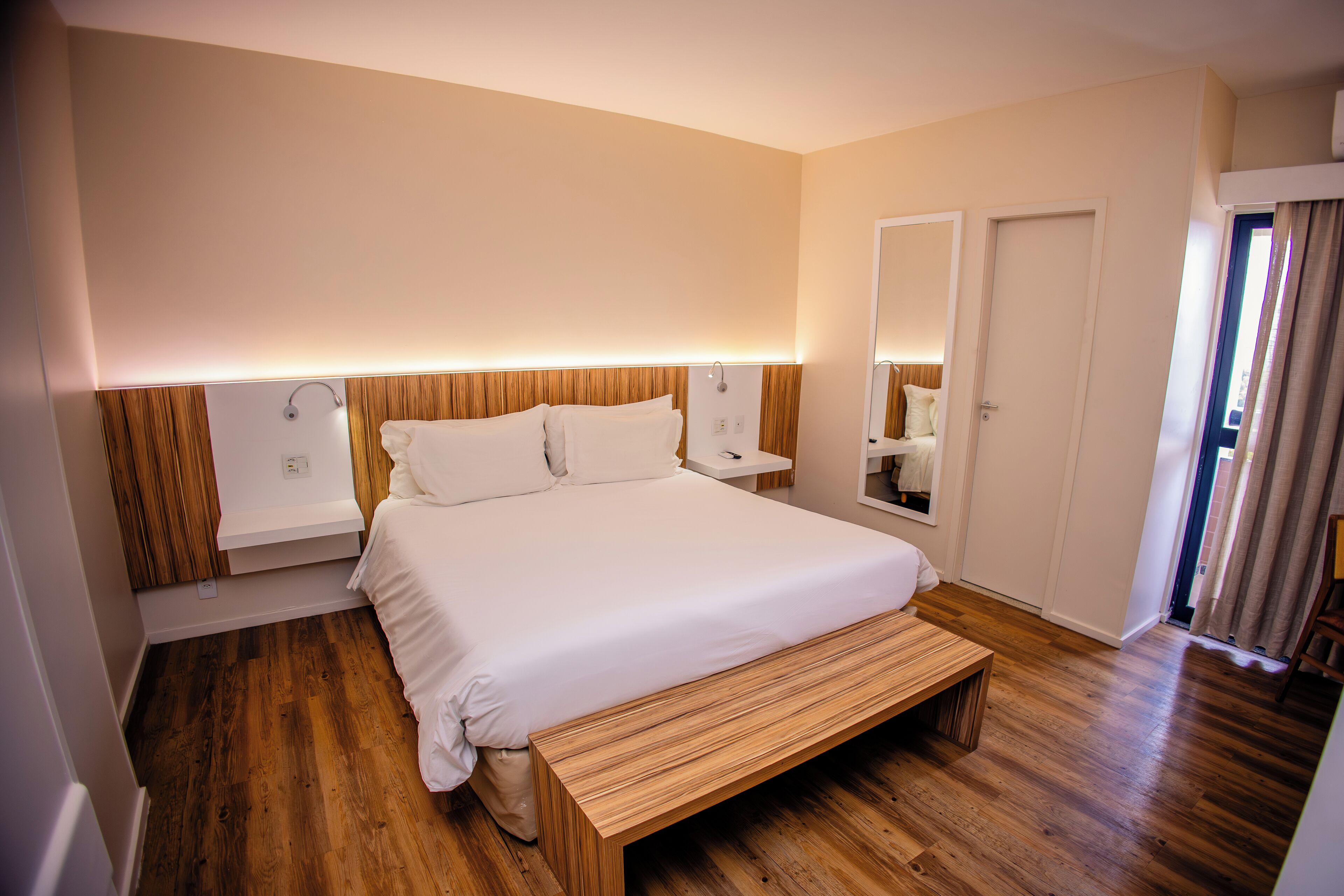 Quarto de hotel moderno com uma cama grande, piso de madeira, um banco ao pé da cama, uma mesa de cabeceira e um espelho na parede.