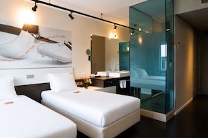 Quarto de hotel moderno com duas camas, um espelho grande, divisória de vidro e uma obra de arte na parede.