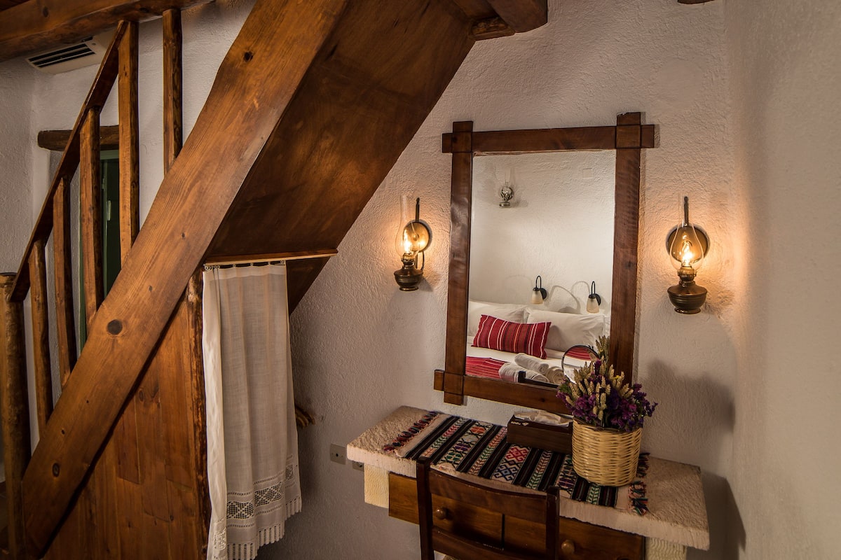 Quarto rústico com escada de madeira, um espelho com castiçais e uma pequena mesa com uma cesta.
