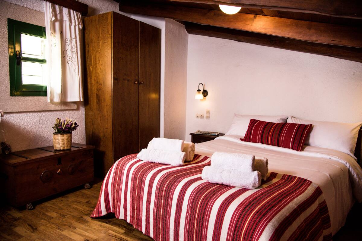 Quarto de hotel com guarda-roupa de madeira, mesa de cabeceira com luminária, cama com roupa de cama listrada e janela com cortinas.