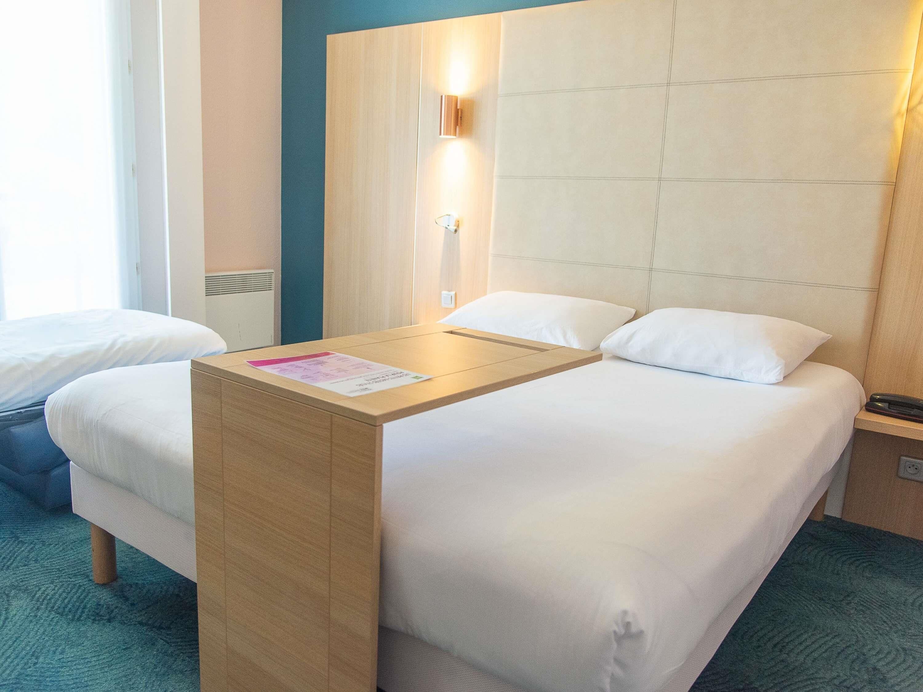 Quarto de hotel com duas camas, uma escrivaninha de madeira, uma mesa de cabeceira e uma luminária fixada na parede.