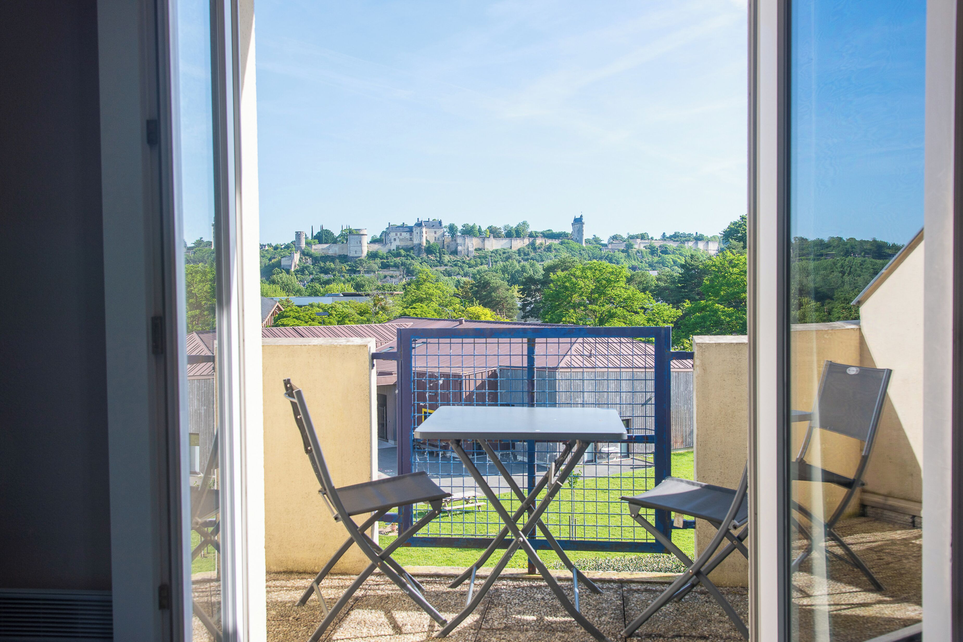 Uma varanda com mesa e cadeiras, vista para uma colina com edifícios e um portão azul.