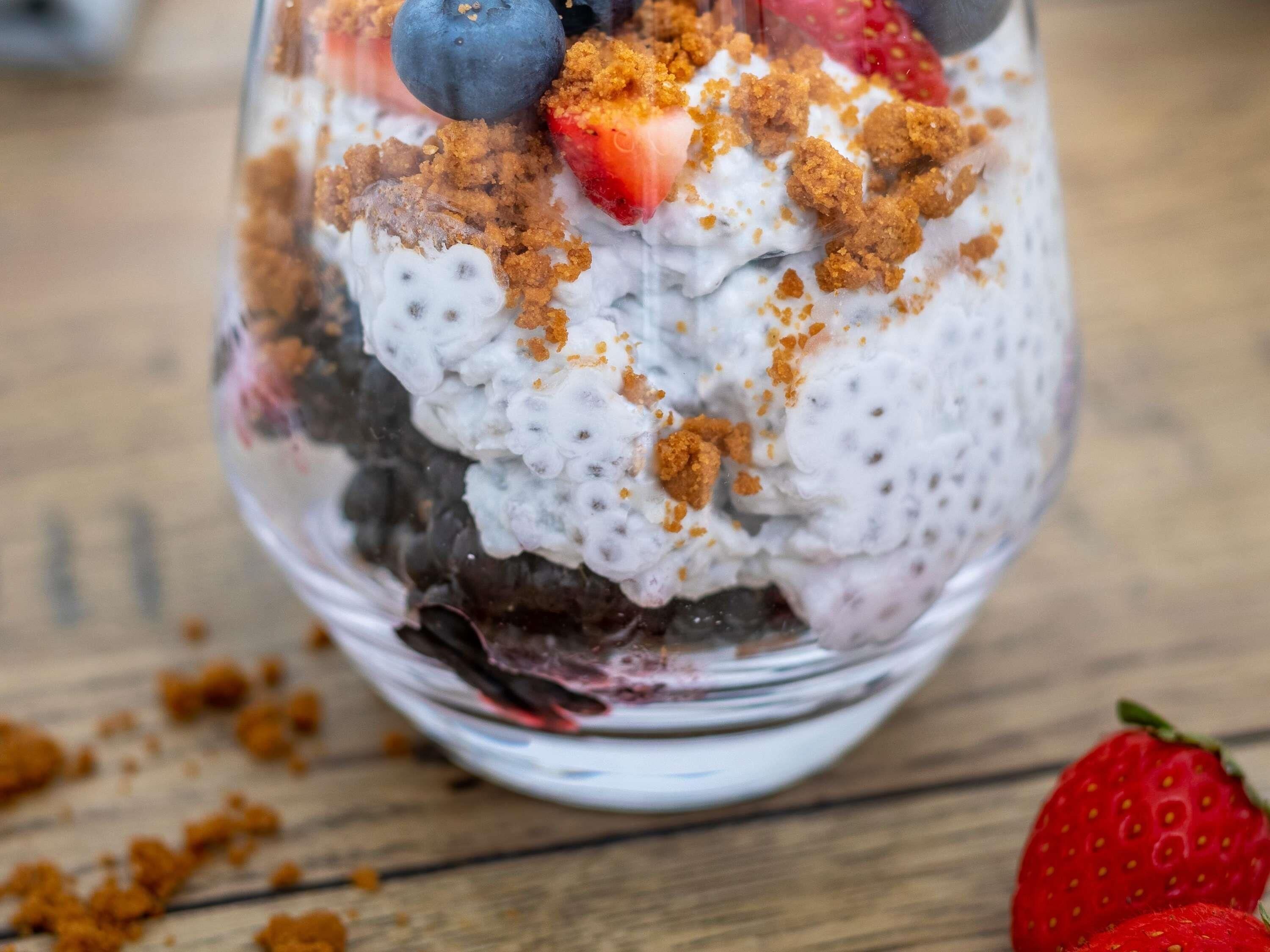 Sobremesa em copo transparente, com camadas de creme branco, migalhas castanhas e frutas vermelhas.