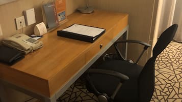 Quarto de hotel com escrivaninha de madeira, cadeira de escritório preta, telefone, luminária e calendário na parede.