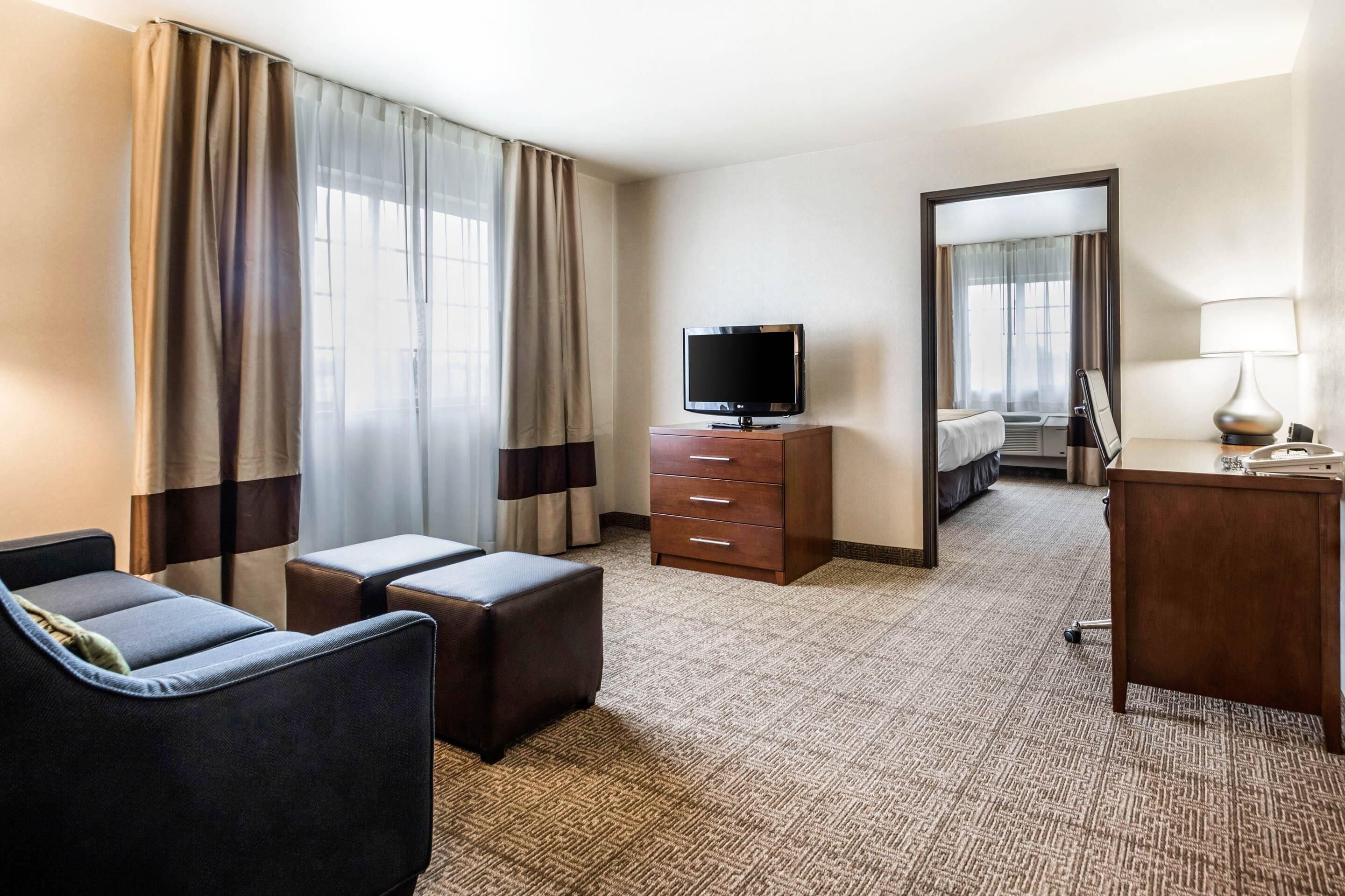 Quarto de hotel com sofá, televisão sobre um aparador de madeira, escrivaninha com luminária e um espelho grande.