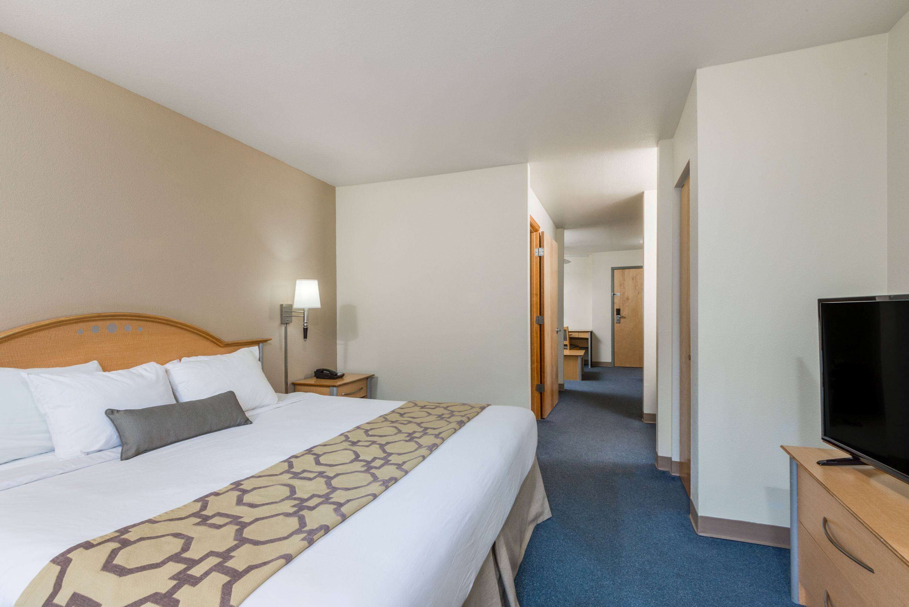 Quarto de hotel com uma cama grande, uma televisão sobre uma cômoda de madeira, e um corredor que leva a outro quarto.