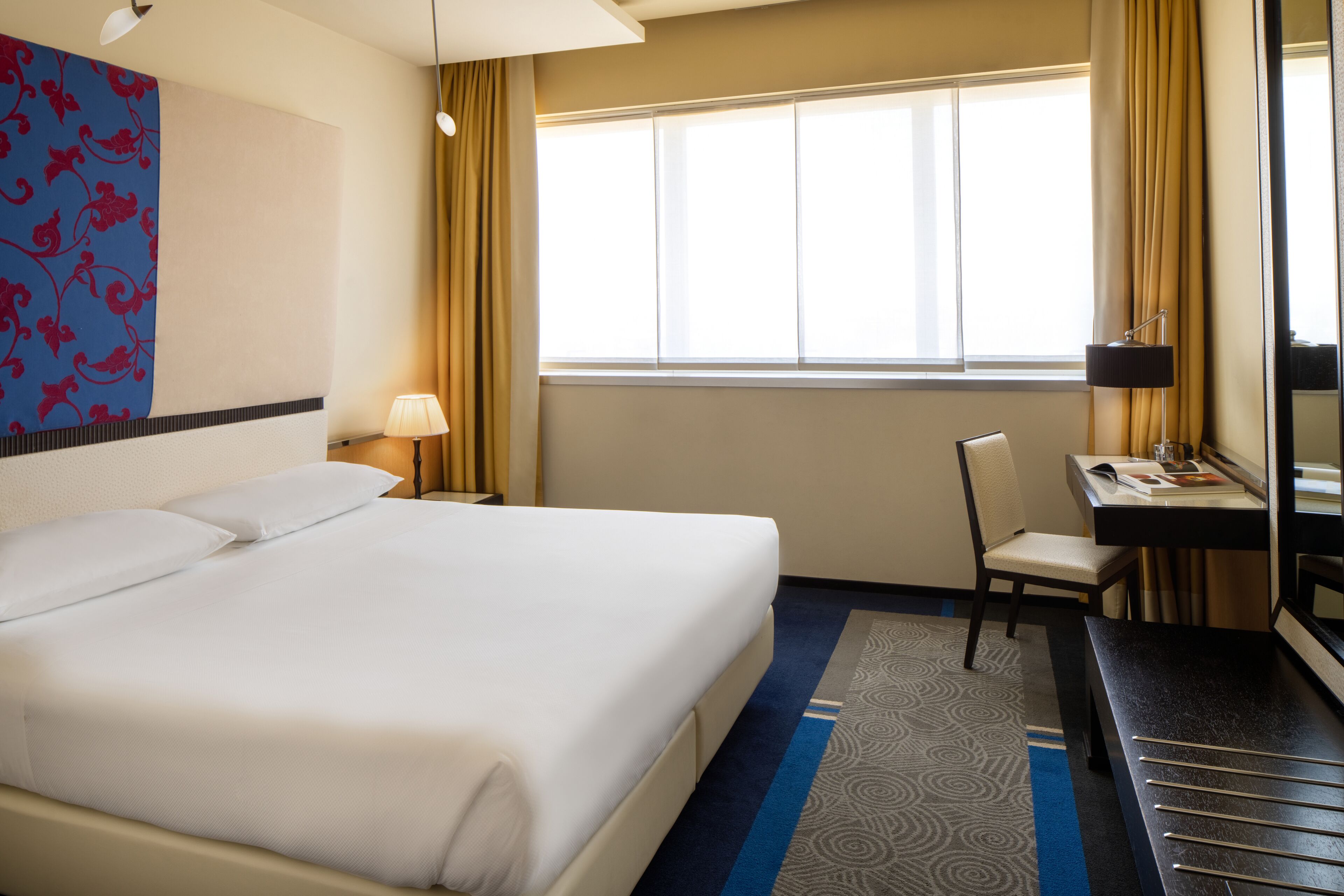 Quarto de hotel com uma cama grande, uma escrivaninha, uma cadeira, um abajur e uma janela com cortinas.