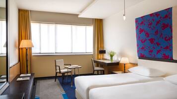 Quarto de hotel com duas camas, uma escrivaninha, uma cadeira, um abajur e uma janela grande com cortinas.