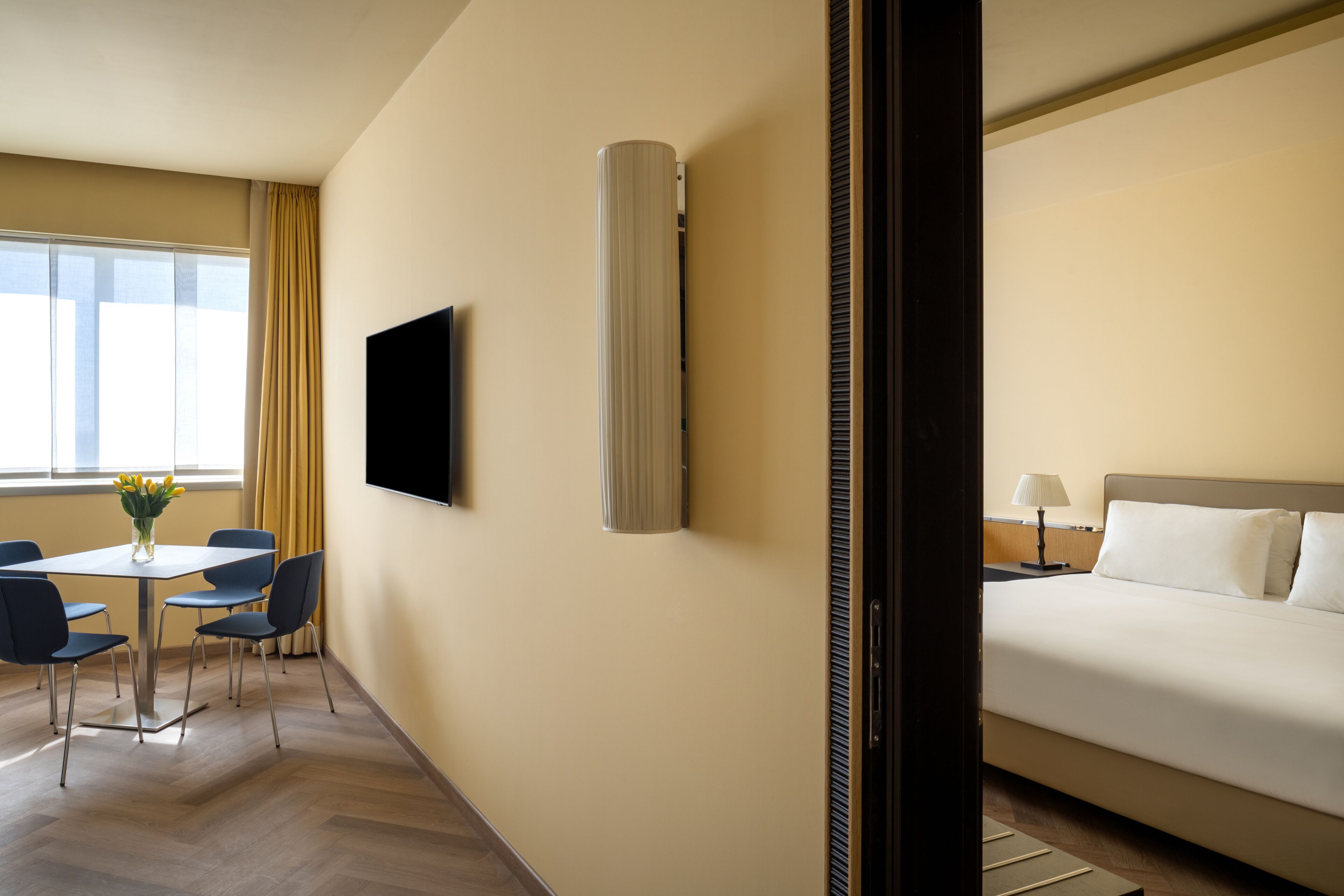 Quarto de hotel com cama, televisão na parede, mesa com cadeiras e janela com cortinas.