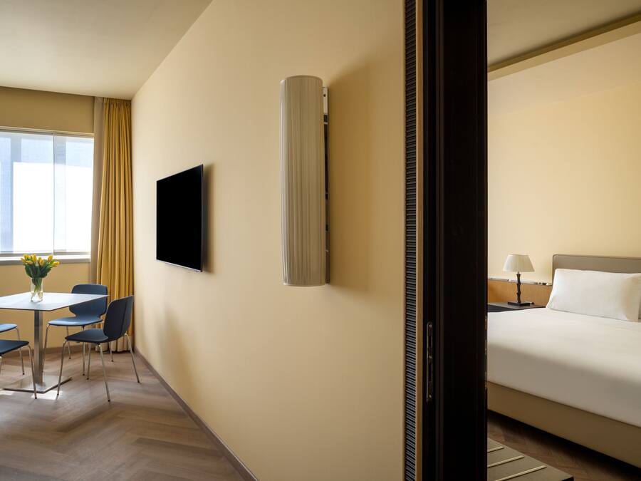 Quarto de hotel com cama, televisão na parede, mesa com cadeiras e janela com cortinas.