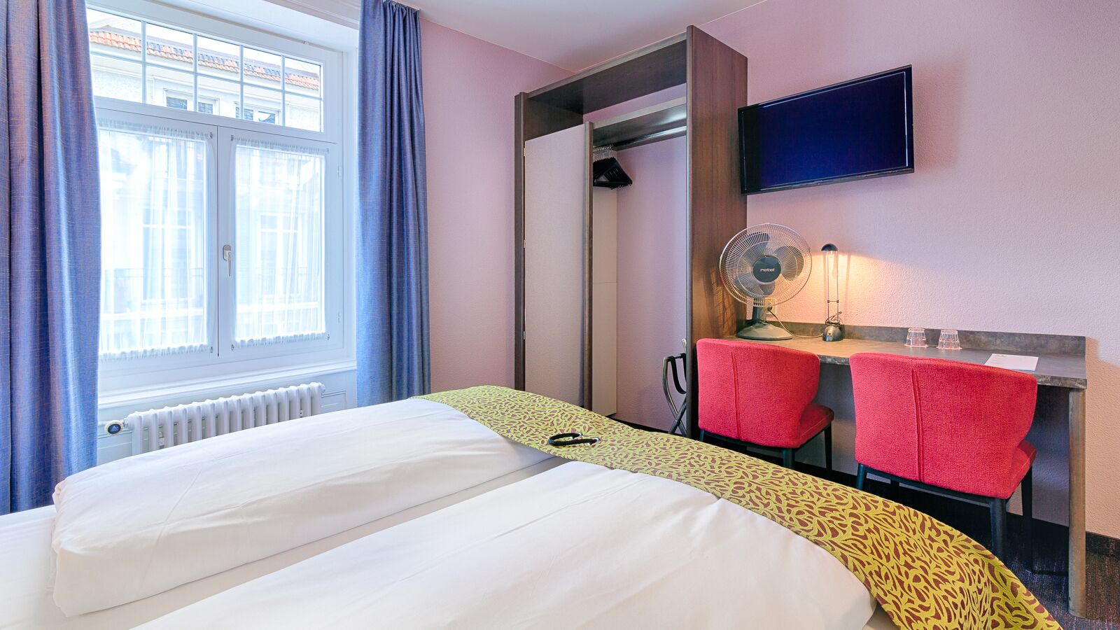 Quarto de hotel com cama, escrivaninha e duas cadeiras vermelhas, televisão e janela com cortinas.