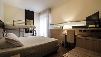 Quarto de hotel com cama, escrivaninha, cadeira, televisão e luminária.