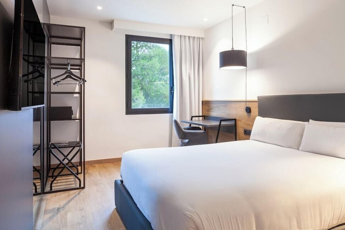 Quarto de hotel com cama, escrivaninha, cadeira, guarda-roupa e janela.
