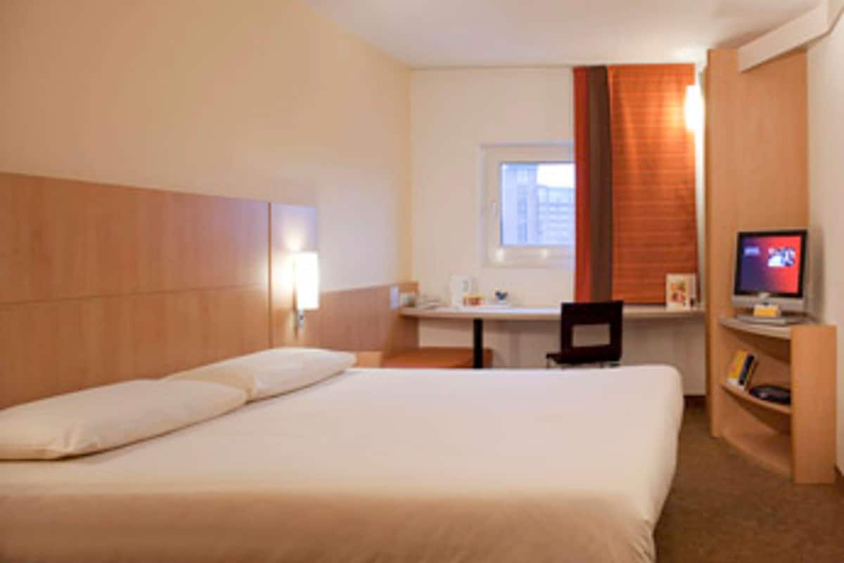Quarto de hotel com cama, escrivaninha, cadeira, televisão e janela com cortinas.