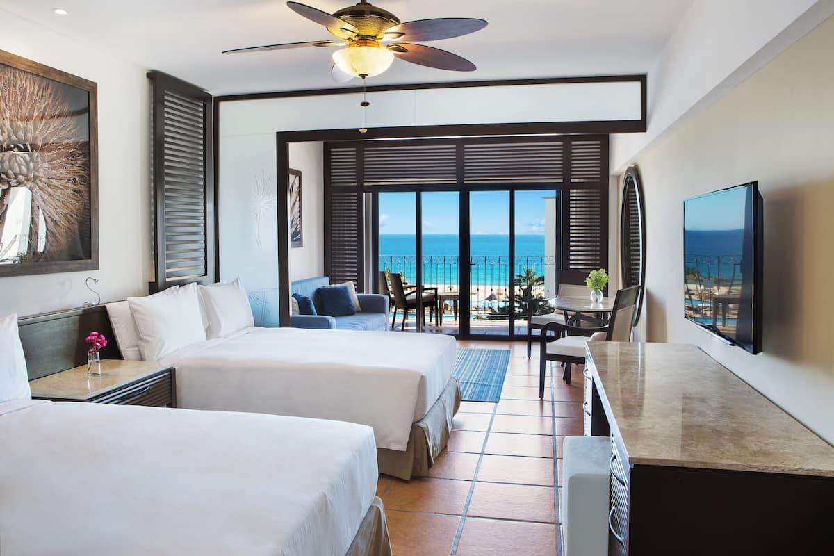 Quarto de hotel com duas camas, varanda com vista para o mar, área de refeições e televisão.