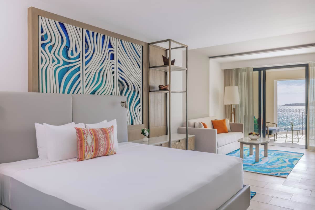 Quarto de hotel moderno com uma cama grande, área de estar com sofá e poltronas, varanda com vista e um painel decorativo na parede.
