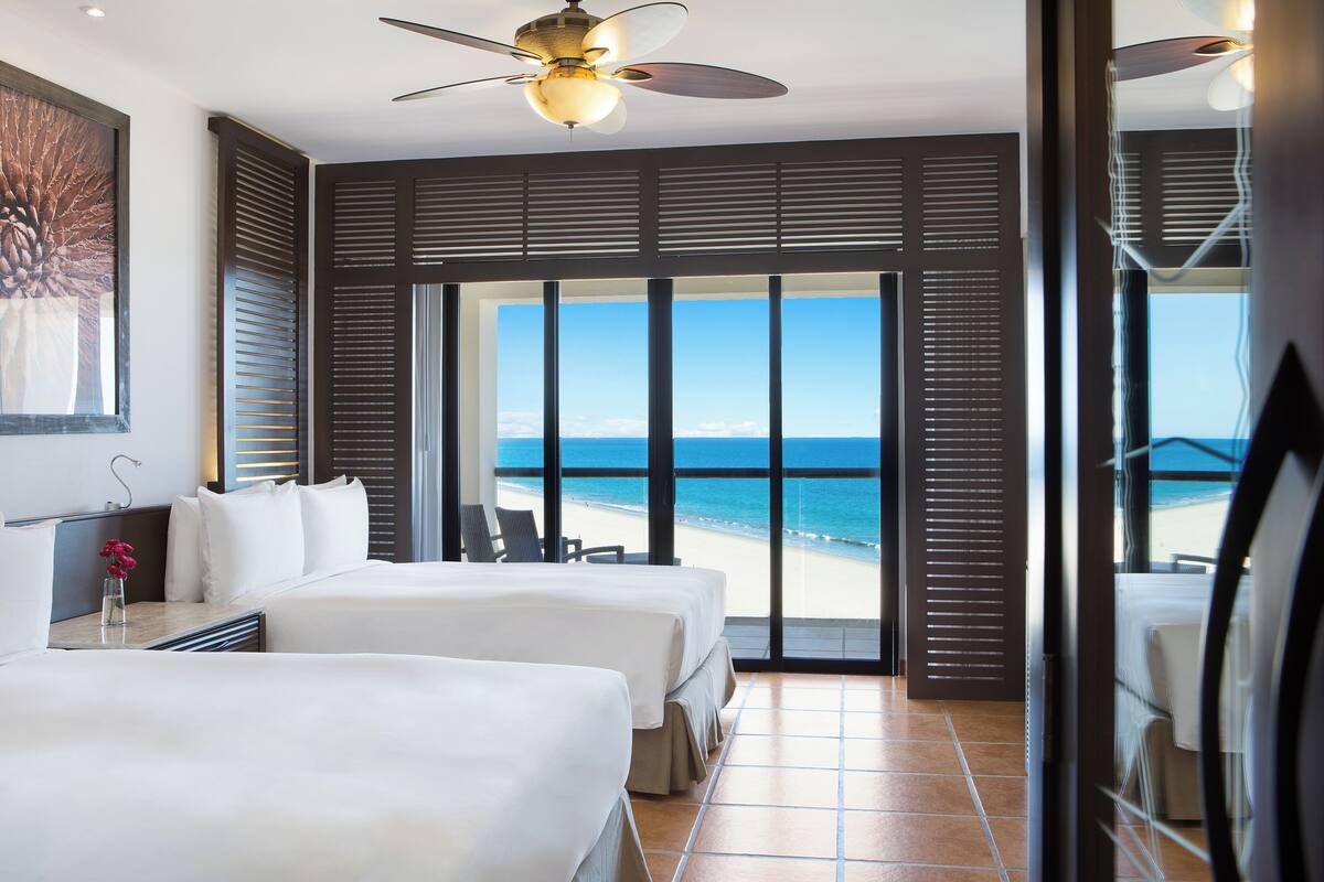 Quarto de hotel com duas camas, ventilador de teto e uma grande janela com vista para a praia.