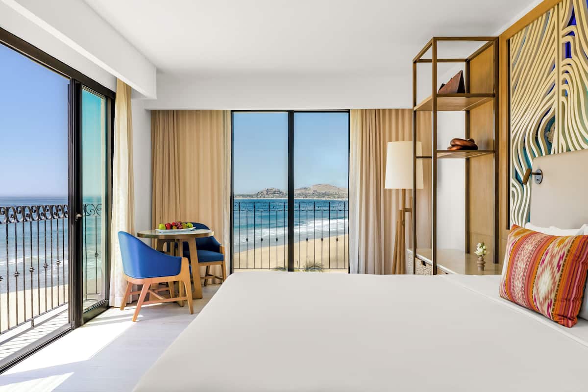 Quarto de hotel com uma cama grande, uma escrivaninha com cadeira, vista para a praia e oceano, e uma varanda com guarda-corpo.