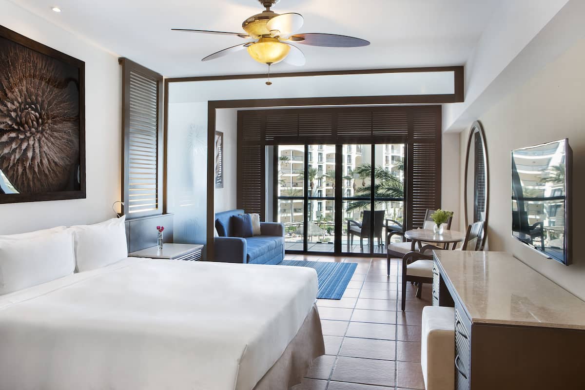 Quarto de hotel com uma cama grande, uma escrivaninha, uma cadeira, um ventilador de teto e vista para o exterior.