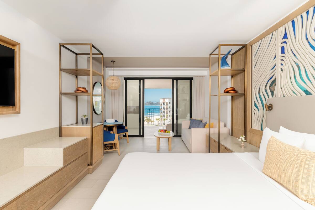 Quarto de hotel moderno com uma cama grande, uma escrivaninha com cadeira, uma mesinha e vista para o mar através de uma porta deslizante.