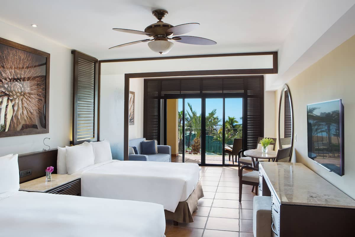 Quarto de hotel com duas camas, um ventilador de teto, uma escrivaninha com cadeira, uma janela grande e vista para palmeiras.