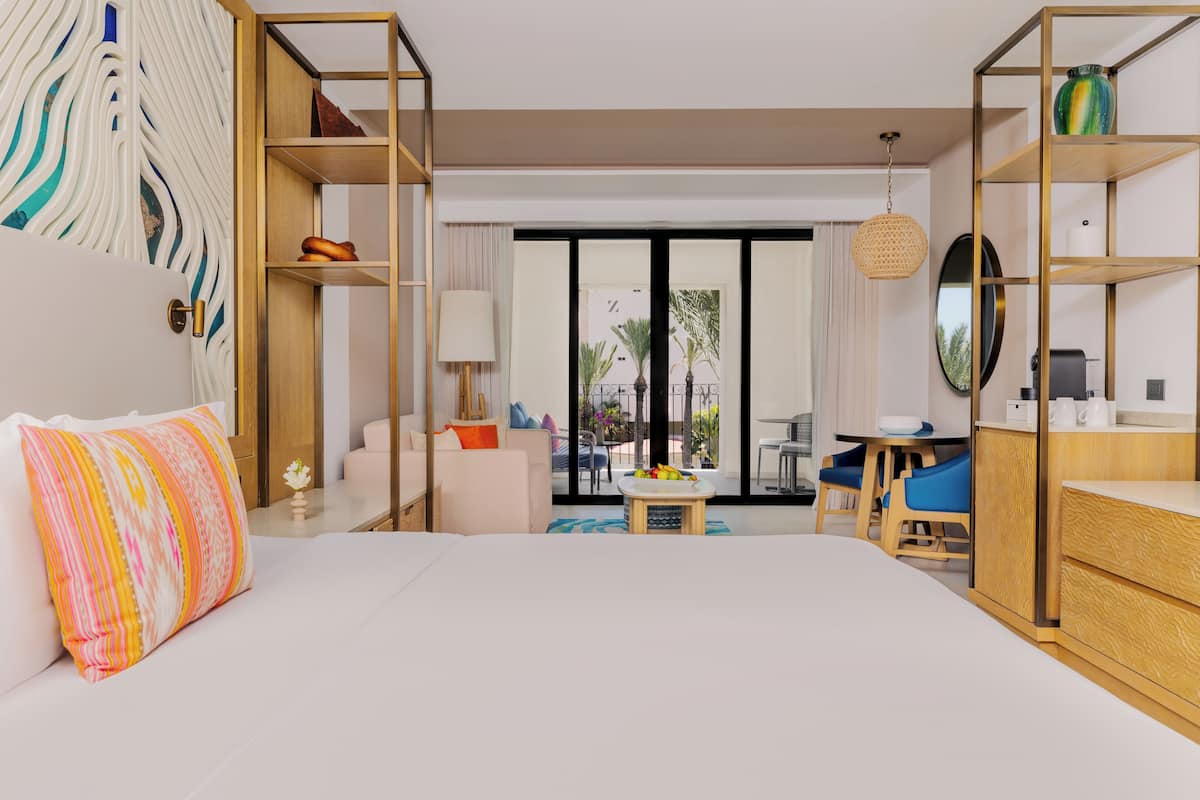Quarto de hotel moderno com uma cama grande, uma escrivaninha, uma cadeira, uma pequena mesa com fruta e vista para o exterior.
