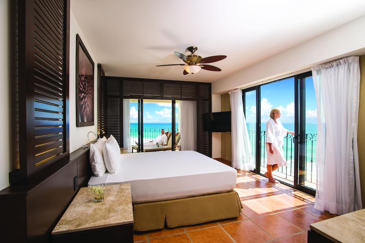 Quarto de hotel com cama grande, ventilador de teto, vista para o oceano e varanda com guarda-corpo.